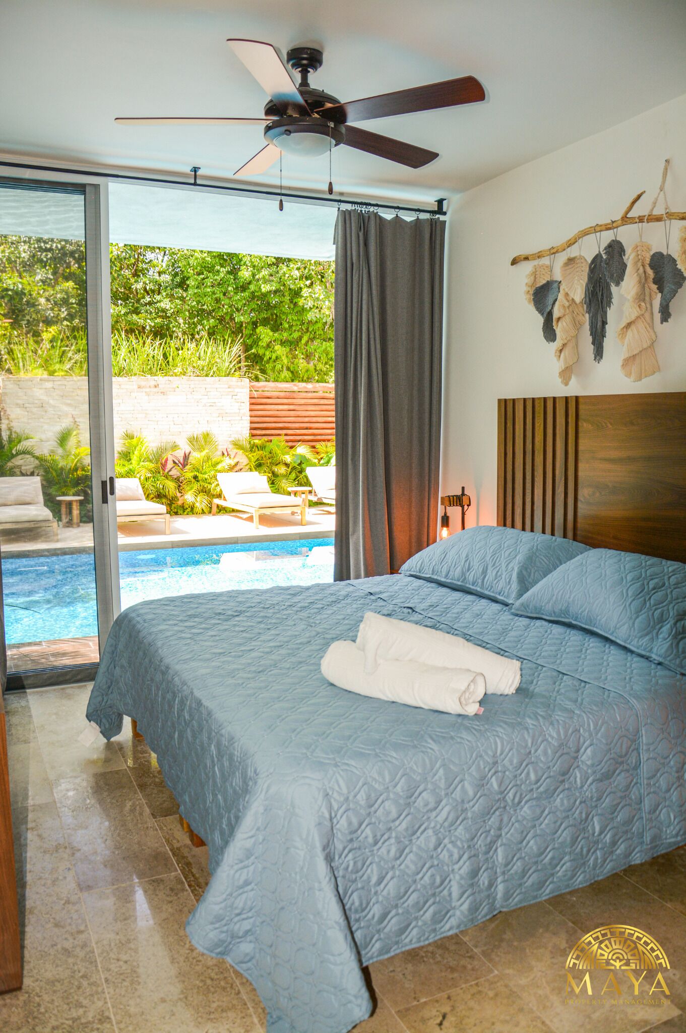 Quarto com cama, ventilador de teto, vista para uma piscina e cabeceira de madeira.