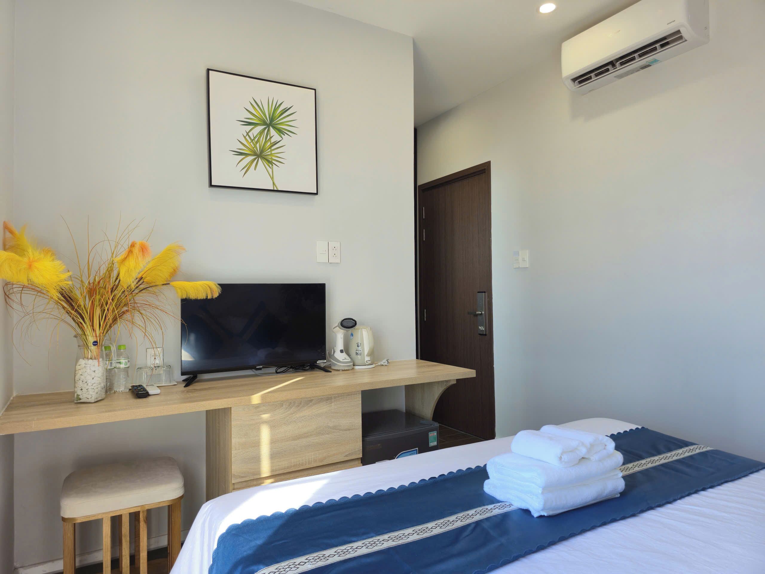 Quarto de hotel com uma cama, uma escrivaninha com uma TV, um vaso com flores amarelas e um quadro na parede.