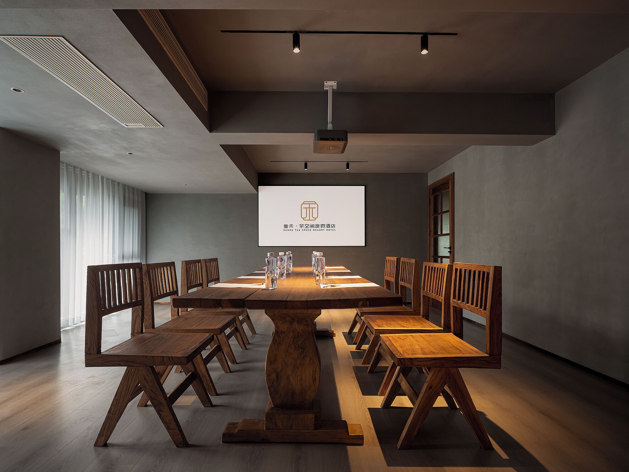 Sala de conferências com uma grande mesa, cadeiras de madeira e uma tela de projetor exibindo o logotipo de um hotel.