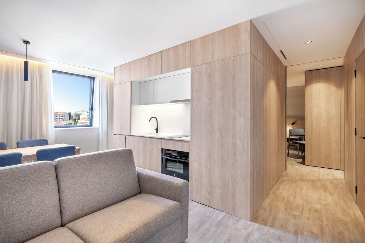 Quarto de hotel moderno com sofá, mesa de jantar com cadeiras, janela grande com cortinas, cozinha compacta com pia e corredor que dá para outro quarto.