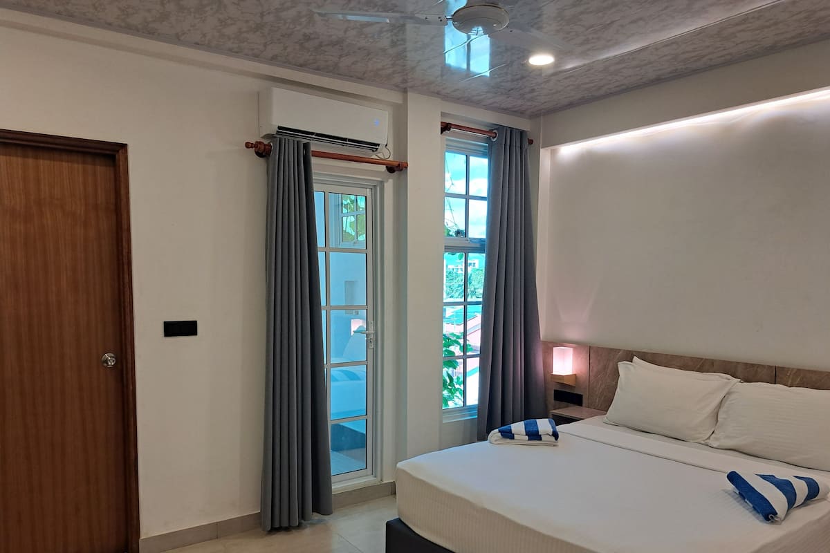 Quarto de hotel com cama, mesa de cabeceira com luminária, janela com cortinas e ar condicionado.