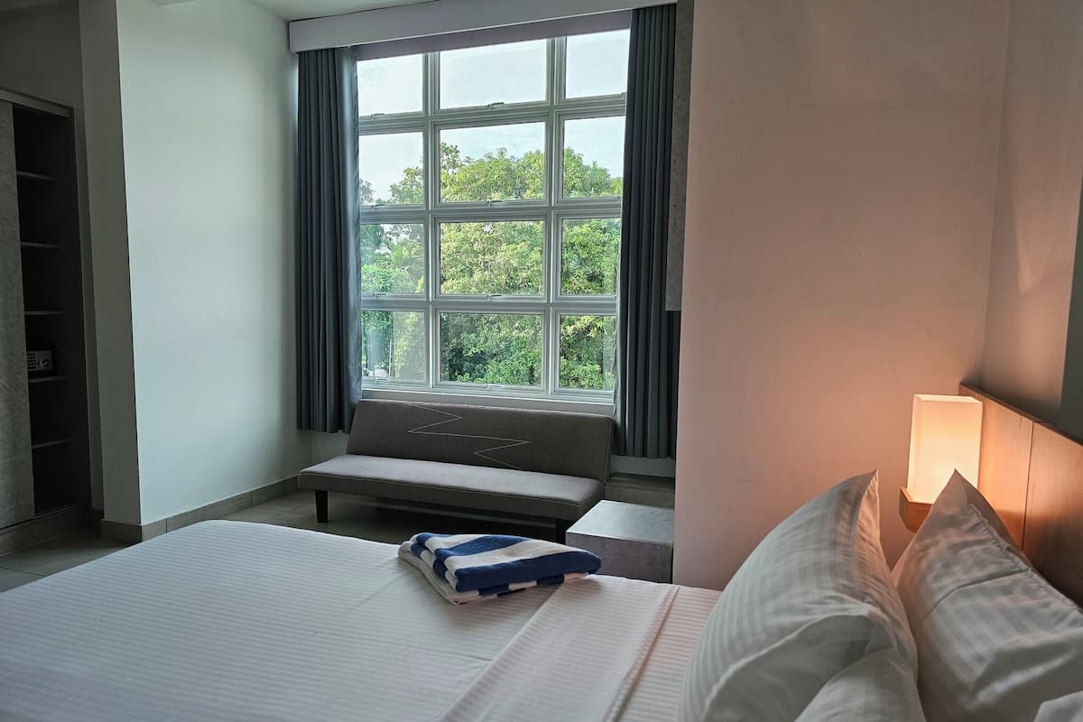Quarto de hotel com uma cama grande, um banquinho perto da janela e vista para o verde lá fora.