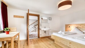 Quarto moderno com piso de madeira, cama com lençóis brancos, cabeceira de madeira, mesa de cabeceira com um vaso vermelho e banheiro com pia e espelho brancos.