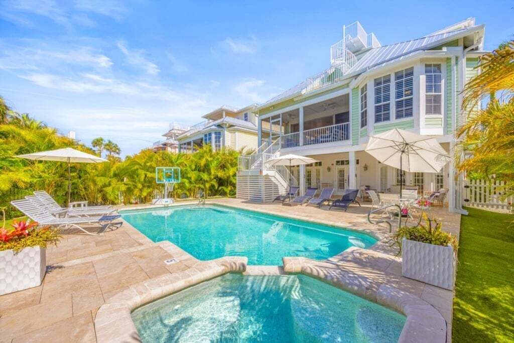 Piscina com espreguiçadeiras e guarda-sóis, rodeada por uma casa com varanda e vista para palmeiras.