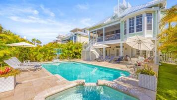 Piscina com espreguiçadeiras e guarda-sóis, rodeada por uma casa com varanda e vista para palmeiras.