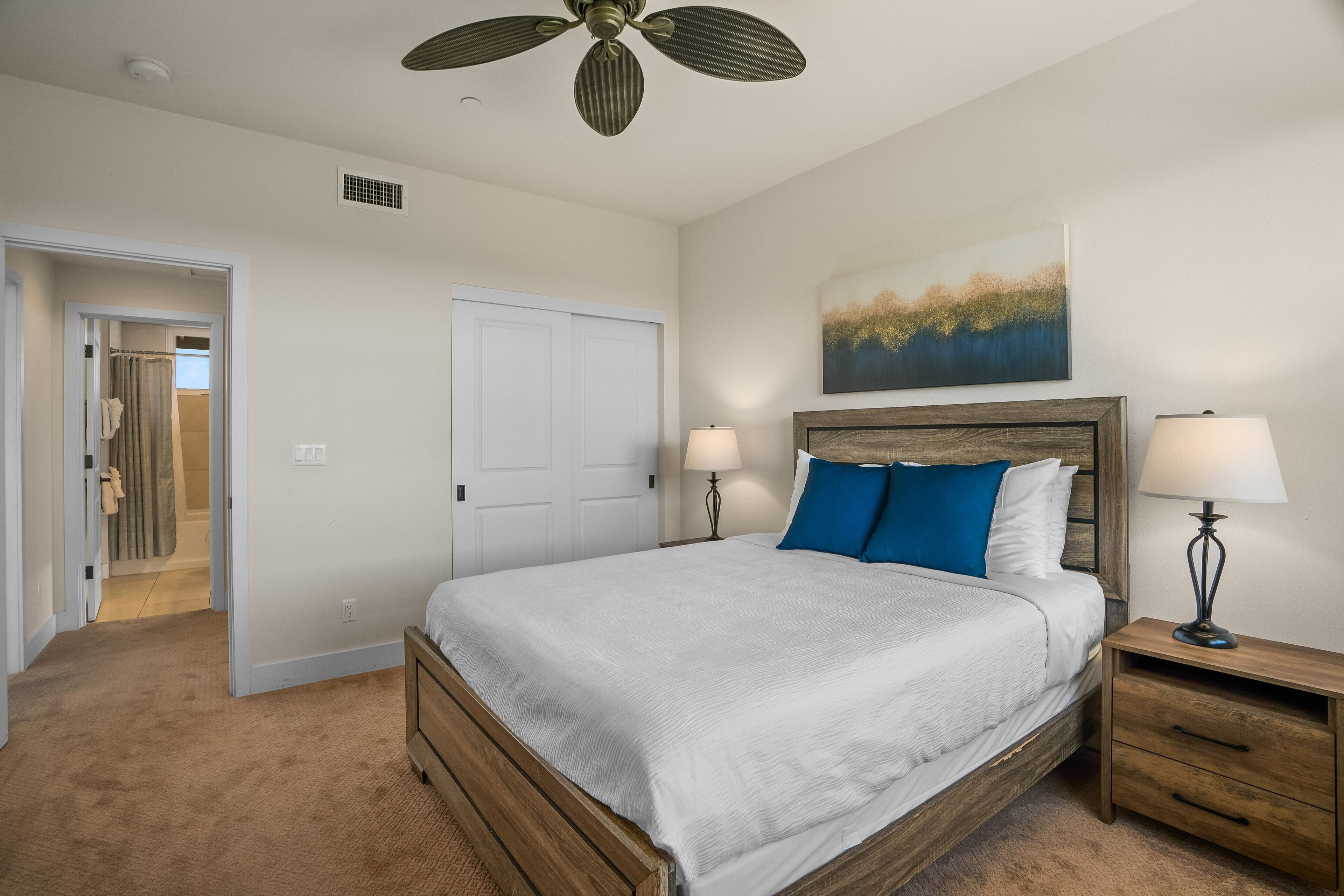 Um quarto com uma cama de madeira, duas luminárias de cabeceira, um ventilador de teto e uma porta que dá para um banheiro.