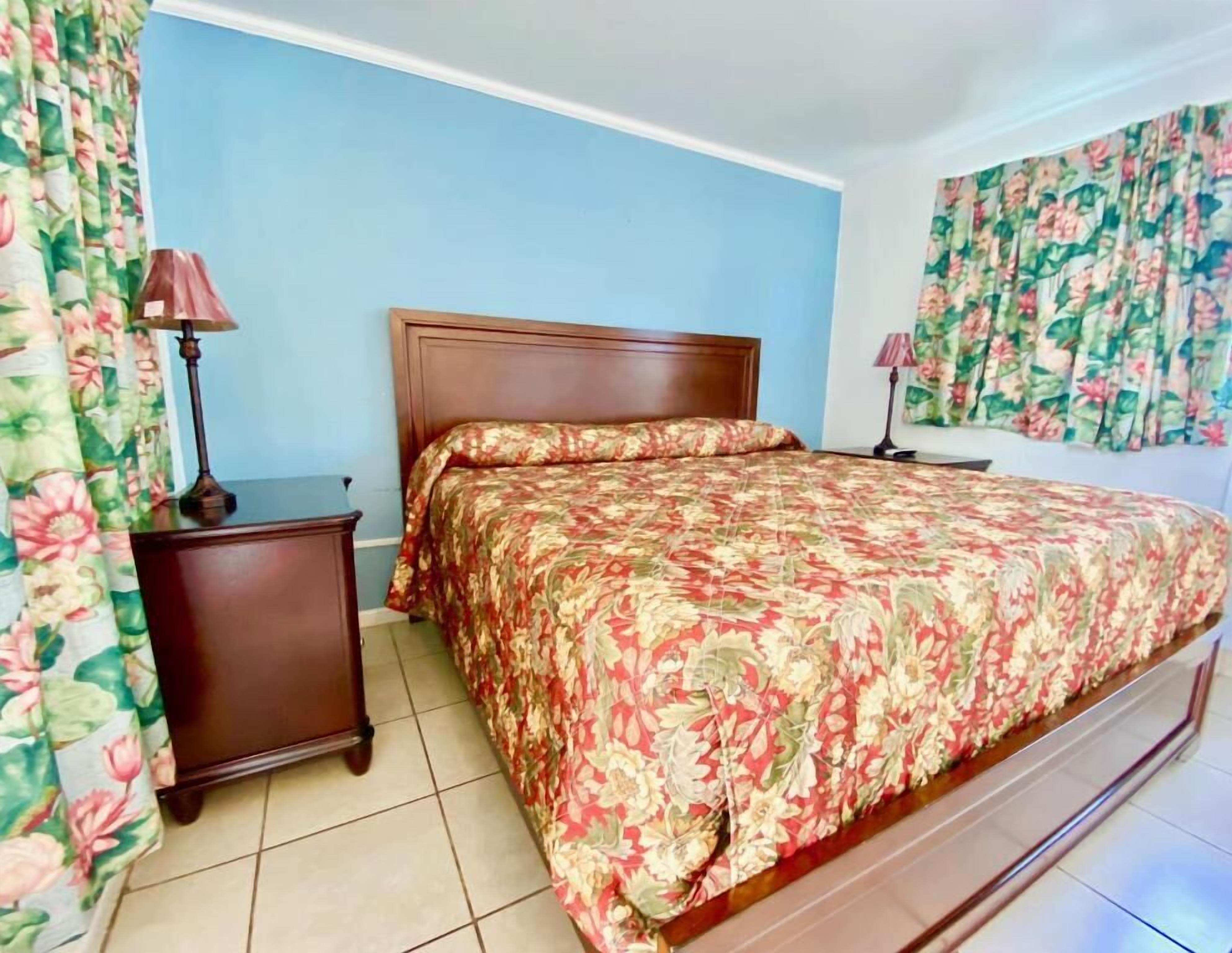 Quarto com cama de madeira, cortinas florais, mesa de cabeceira e luminária.