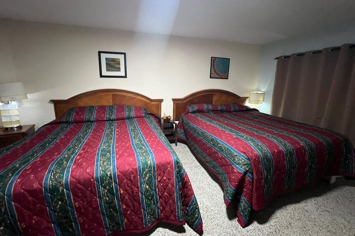Duas camas de solteiro com roupa de cama listrada em vermelho e verde, uma mesa de cabeceira com abajur, e duas fotografias emolduradas na parede.