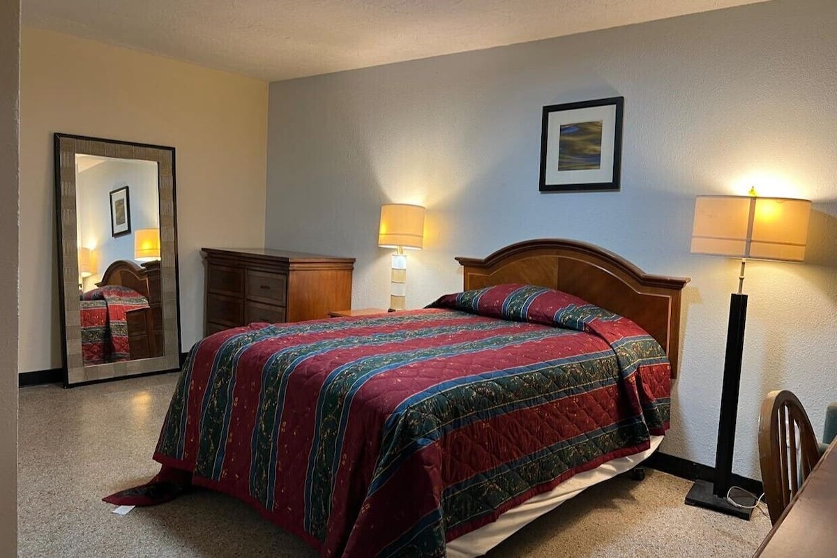 Uma cama bem arrumada com colcha estampada, cabeceira de madeira, duas luminárias de cabeceira, um espelho e um quadro na parede.