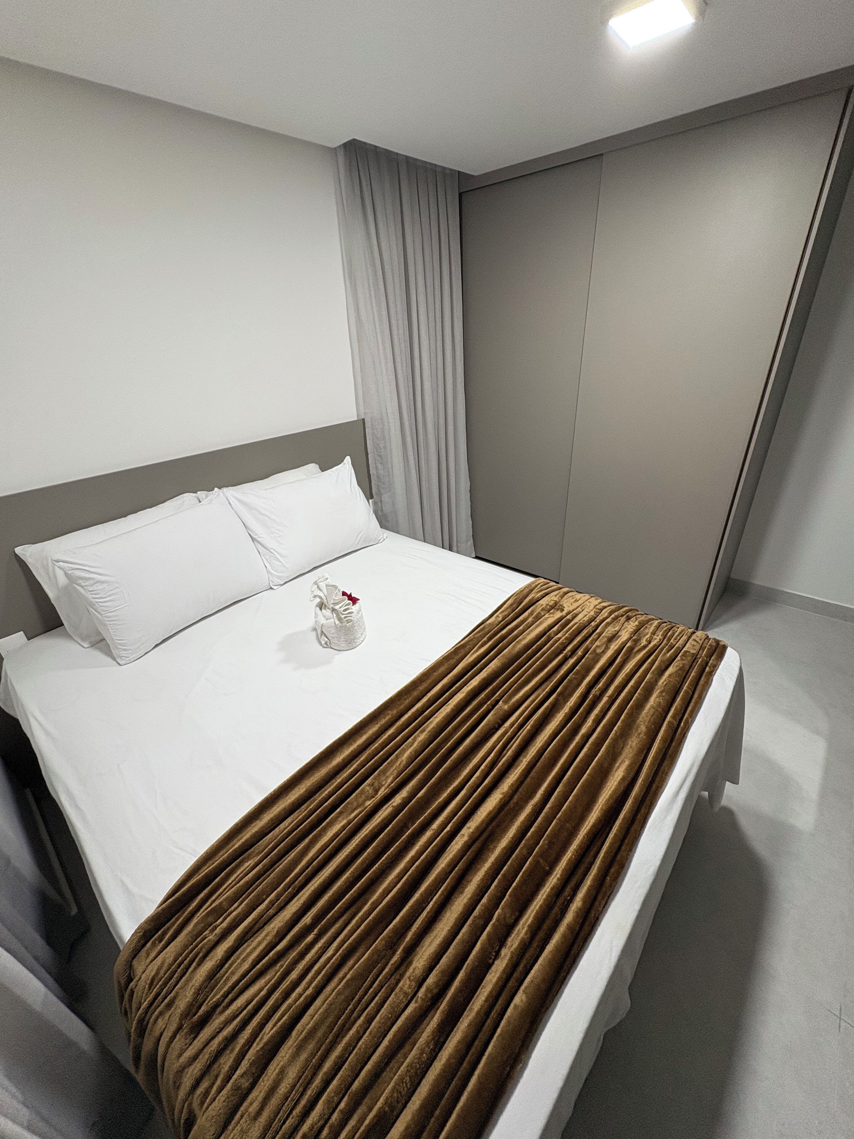 Quarto de hotel moderno com cama bem arrumada, manta marrom e um pequeno objeto decorativo no travesseiro.
