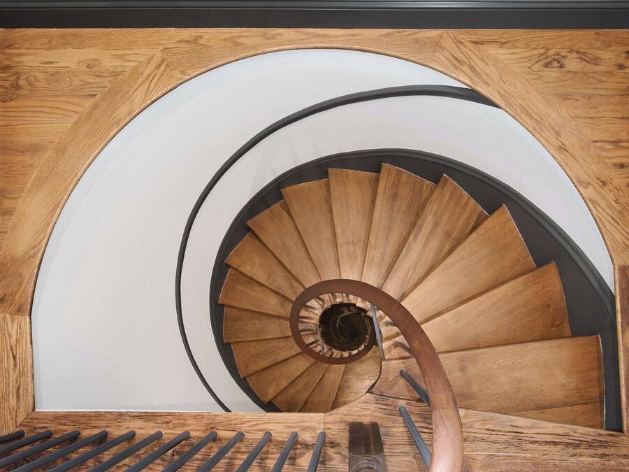 Escadaria em espiral com degraus de madeira e corrimão de metal, envolvida por uma estrutura de madeira e paredes brancas.