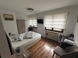 Quarto com cama, escrivaninha, cadeira, radiador, televisão e janela com persianas.
