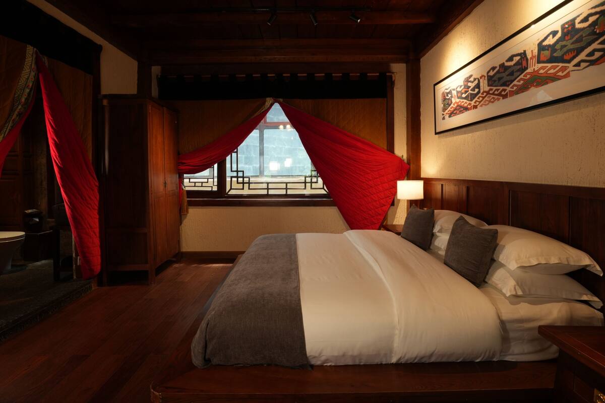 Quarto de hotel com uma cama grande, piso de madeira, janela com cortinas vermelhas e um quadro na parede.