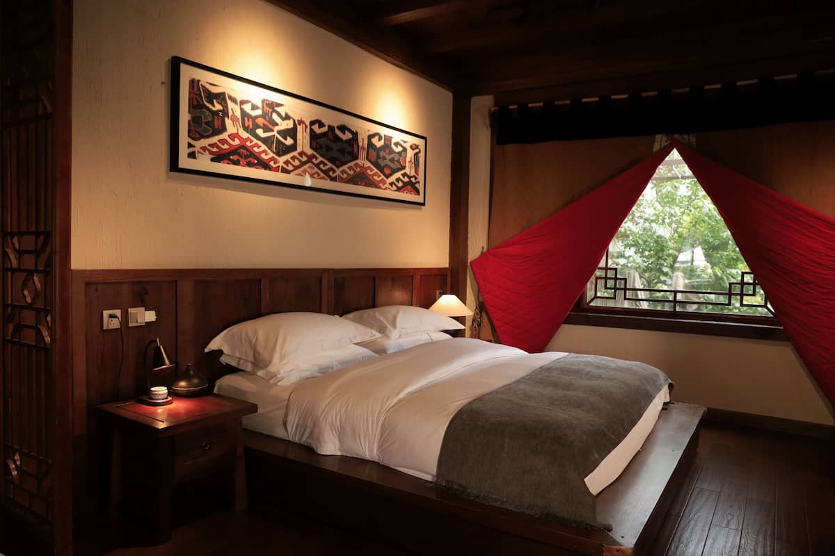 Quarto com cama, mesa de cabeceira, luminária, janela com cortina vermelha e um quadro pendurado na parede.