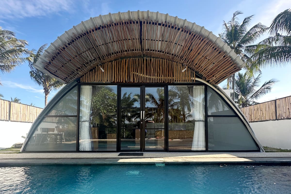 Estrutura moderna à beira da piscina, com teto curvo e grandes janelas, cercada por uma piscina azul límpida e palmeiras.