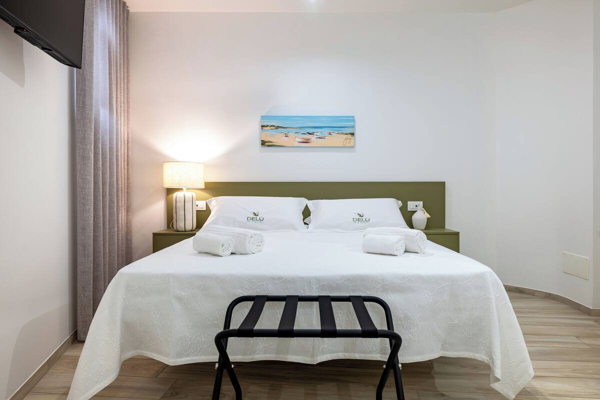 Uma cama bem arrumada com lençóis brancos, um quadro com uma paisagem de praia e um abajur.