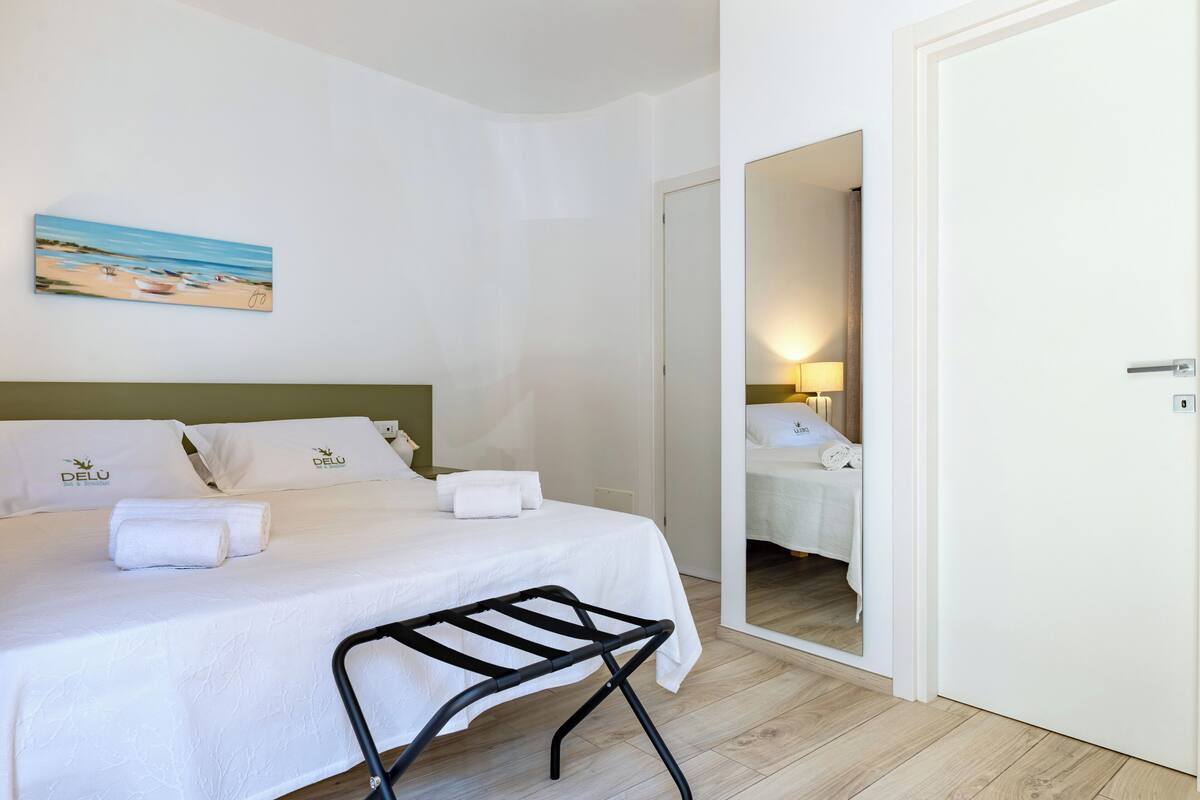 Quarto de hotel com uma cama, uma pintura de uma praia, um espelho e uma porta que dá para outro quarto.