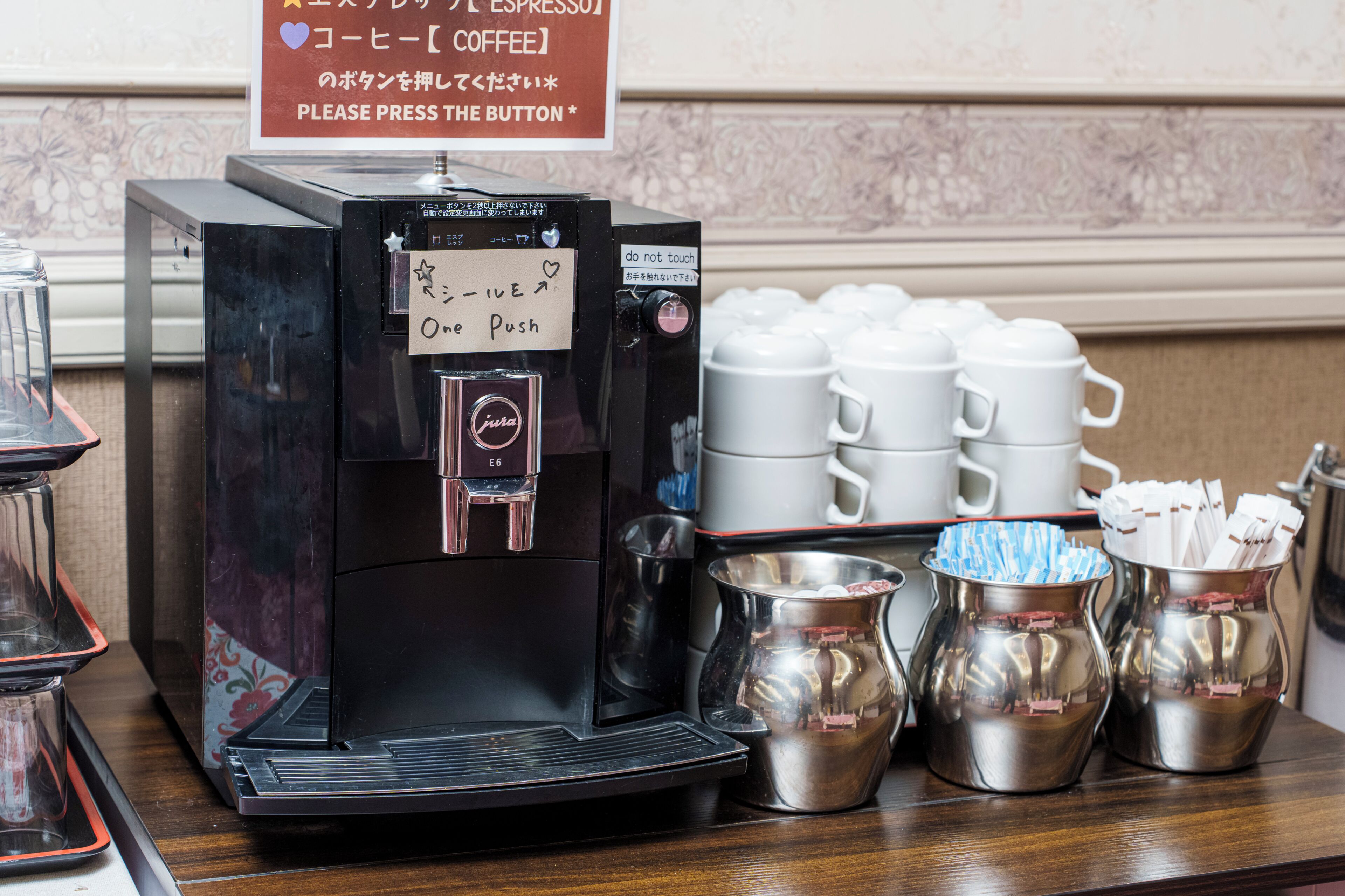 Máquina de café com placa indicando para pressionar o botão, xícaras de café e recipientes de aço inoxidável com cápsulas de café.