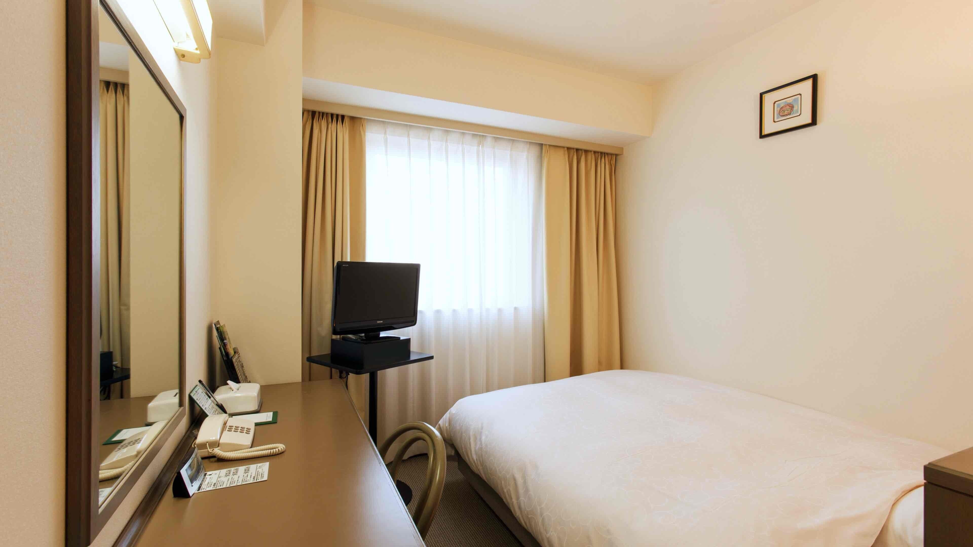 Quarto de hotel com cama, escrivaninha com telefone e televisão, e janela com cortinas.