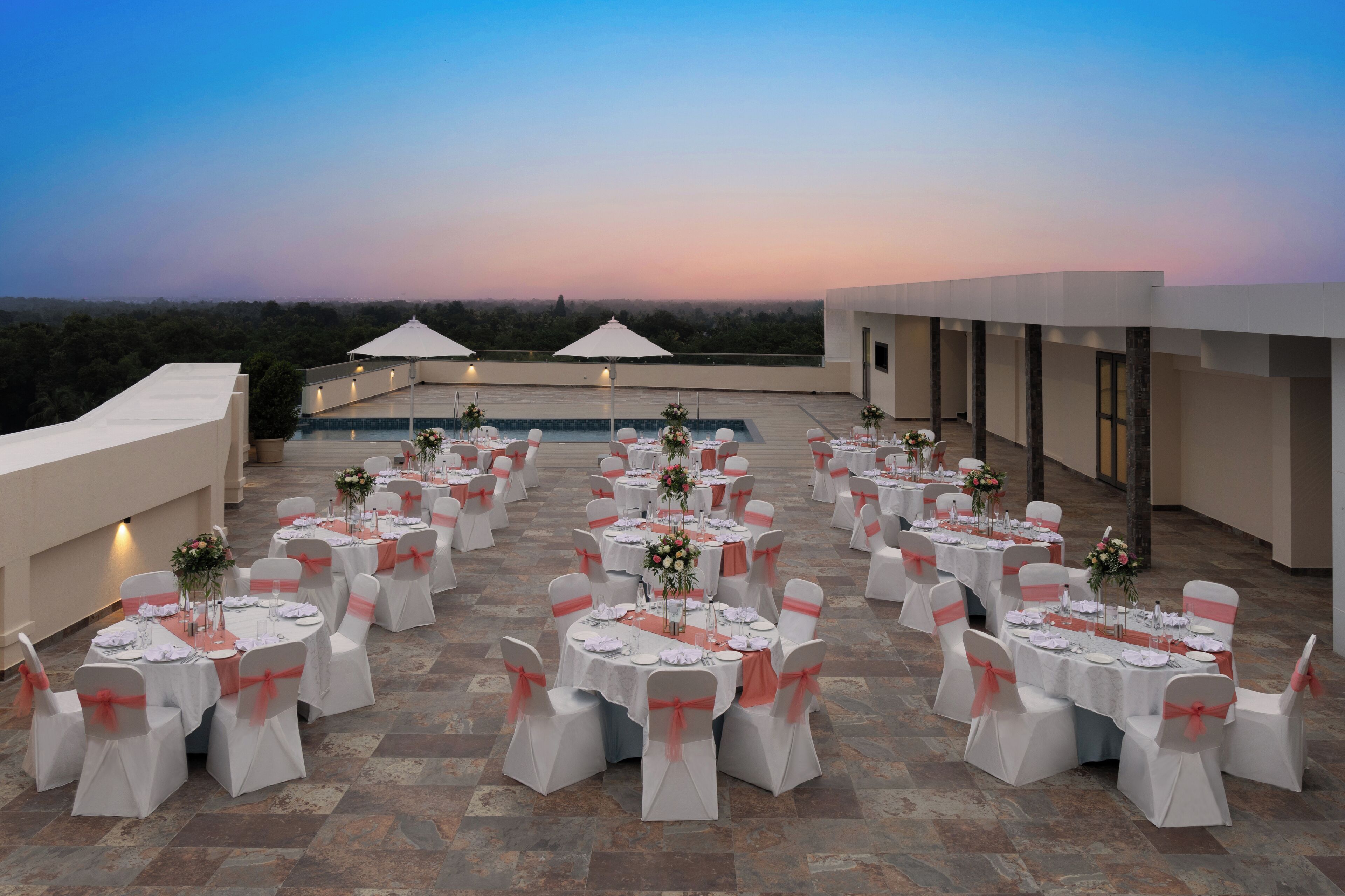Uma área de refeições no terraço, com mesas e cadeiras redondas, cada uma decorada com toalhas brancas e fitas cor-de-rosa, com o céu crepuscular ao fundo.