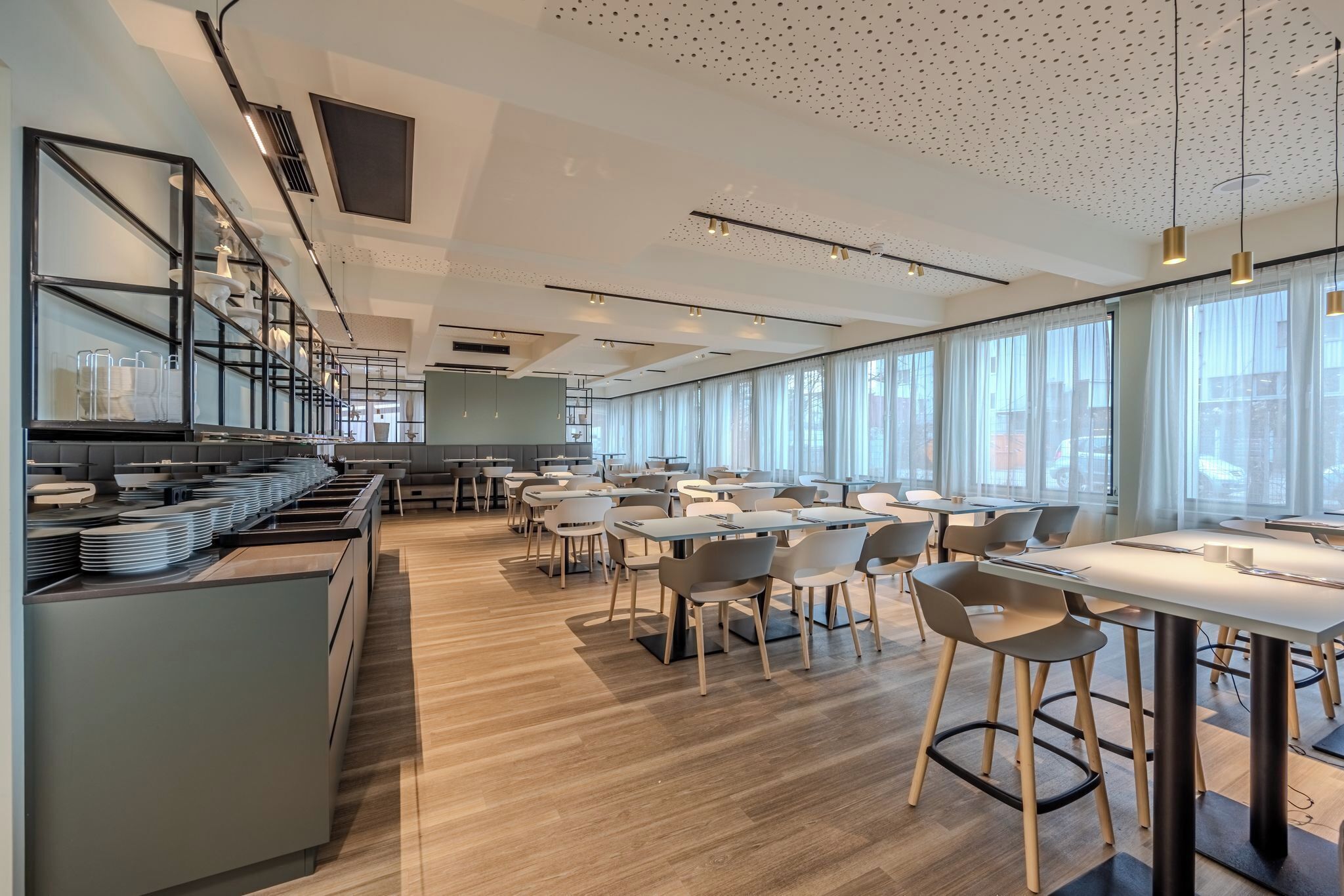 Uma área de jantar moderna com mesas e cadeiras compridas, uma bancada de cozinha com louças e grandes janelas que proporcionam luz natural.