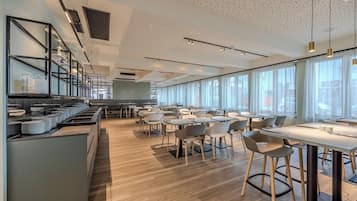 Uma área de jantar moderna com mesas e cadeiras compridas, uma bancada de cozinha com louças e grandes janelas que proporcionam luz natural.