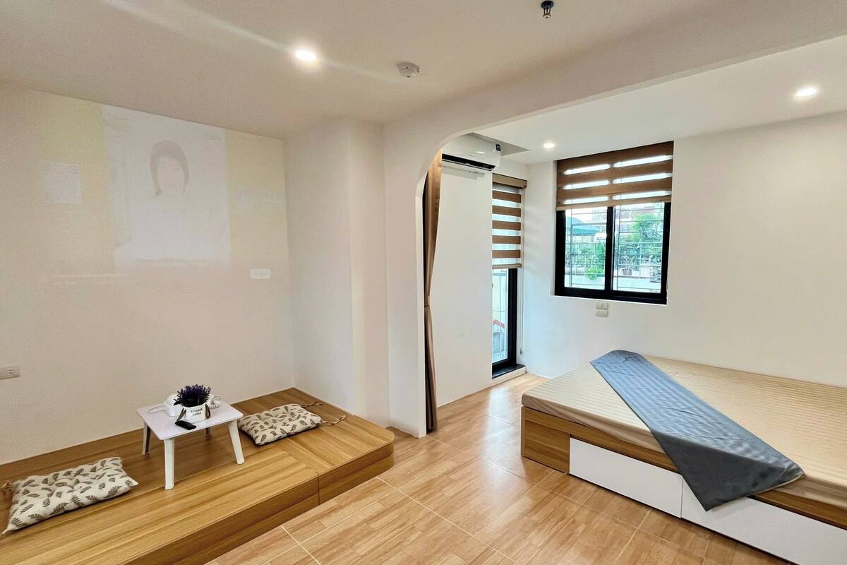 Quarto compacto com cama, mesa pequena com abajur, ar condicionado na parede e janela com persianas.