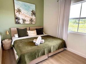 Quarto com uma cama, um quadro de uma cena de praia, uma janela com vista e um abajur.