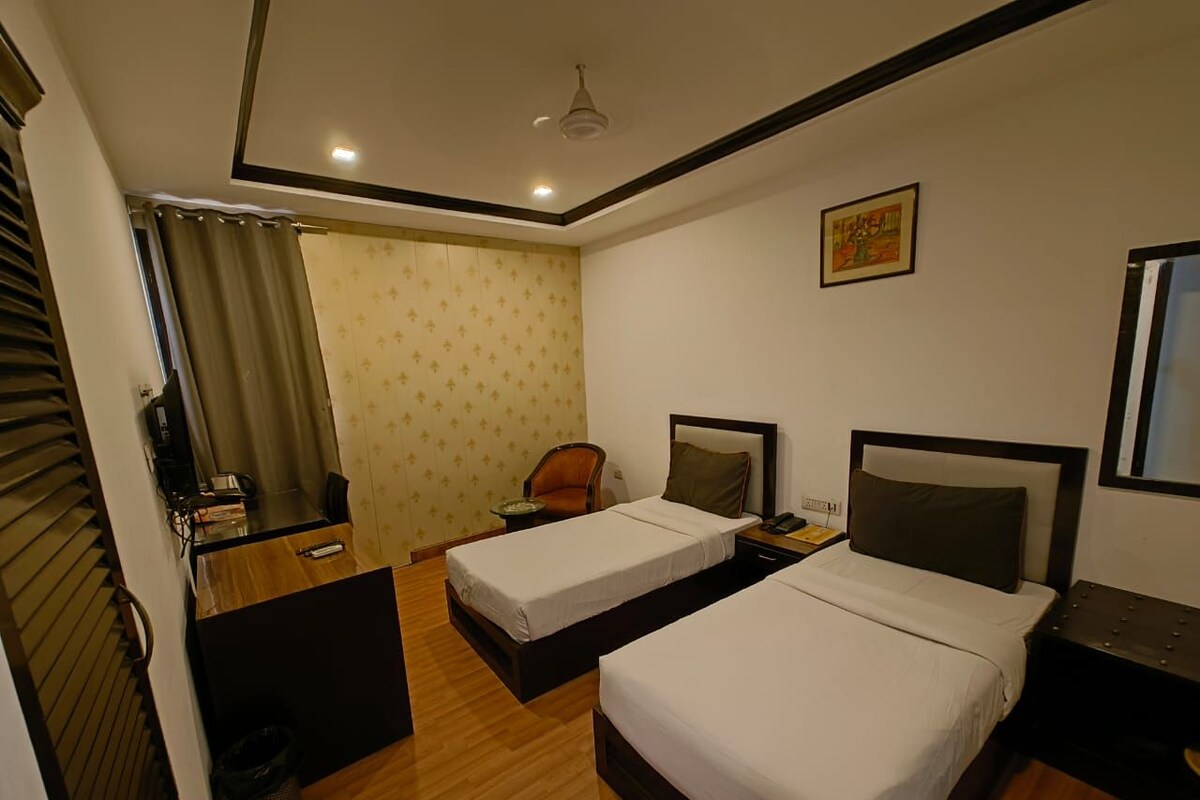 Quarto de hotel com duas camas, uma escrivaninha, uma cadeira, uma mesinha e um ventilador de teto.