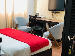 Quarto de hotel com uma cama, duas cadeiras, uma escrivaninha, uma televisão e uma mesinha.