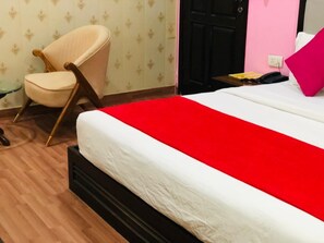 Uma cama com um cobertor vermelho, uma cadeira, uma mesinha de cabeceira com um telefone e uma porta ao fundo.