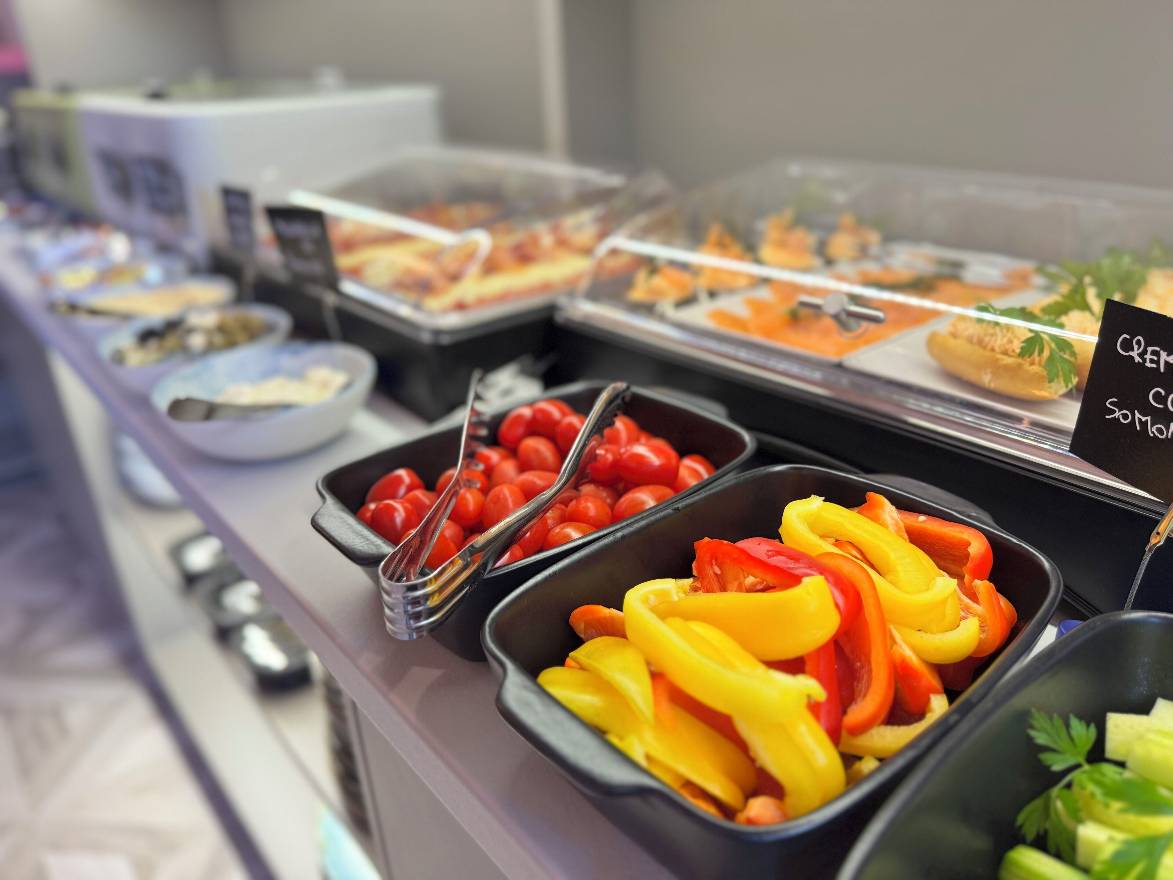 Buffet com diversas opções de comida, incluindo tomates, pimentões e um aviso indicando sanduíches de cream cheese e pepino.
