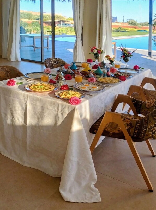 Mesa de jantar posta com diversos pratos, vista para piscina e ambiente externo ensolarado.
