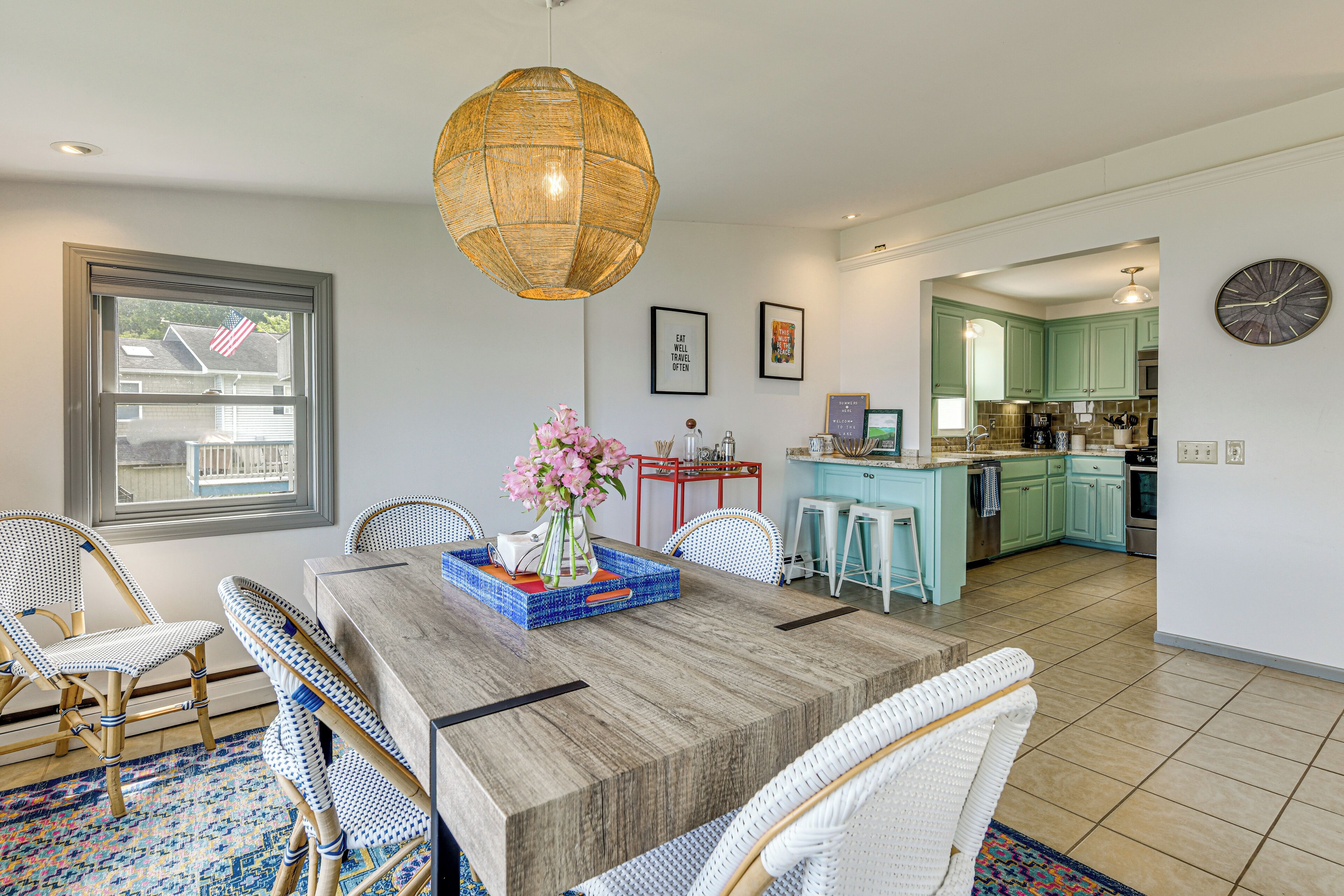 Uma sala de jantar com mesa de madeira, cadeiras de vime, luminária pendente e vista para a cozinha ao fundo.