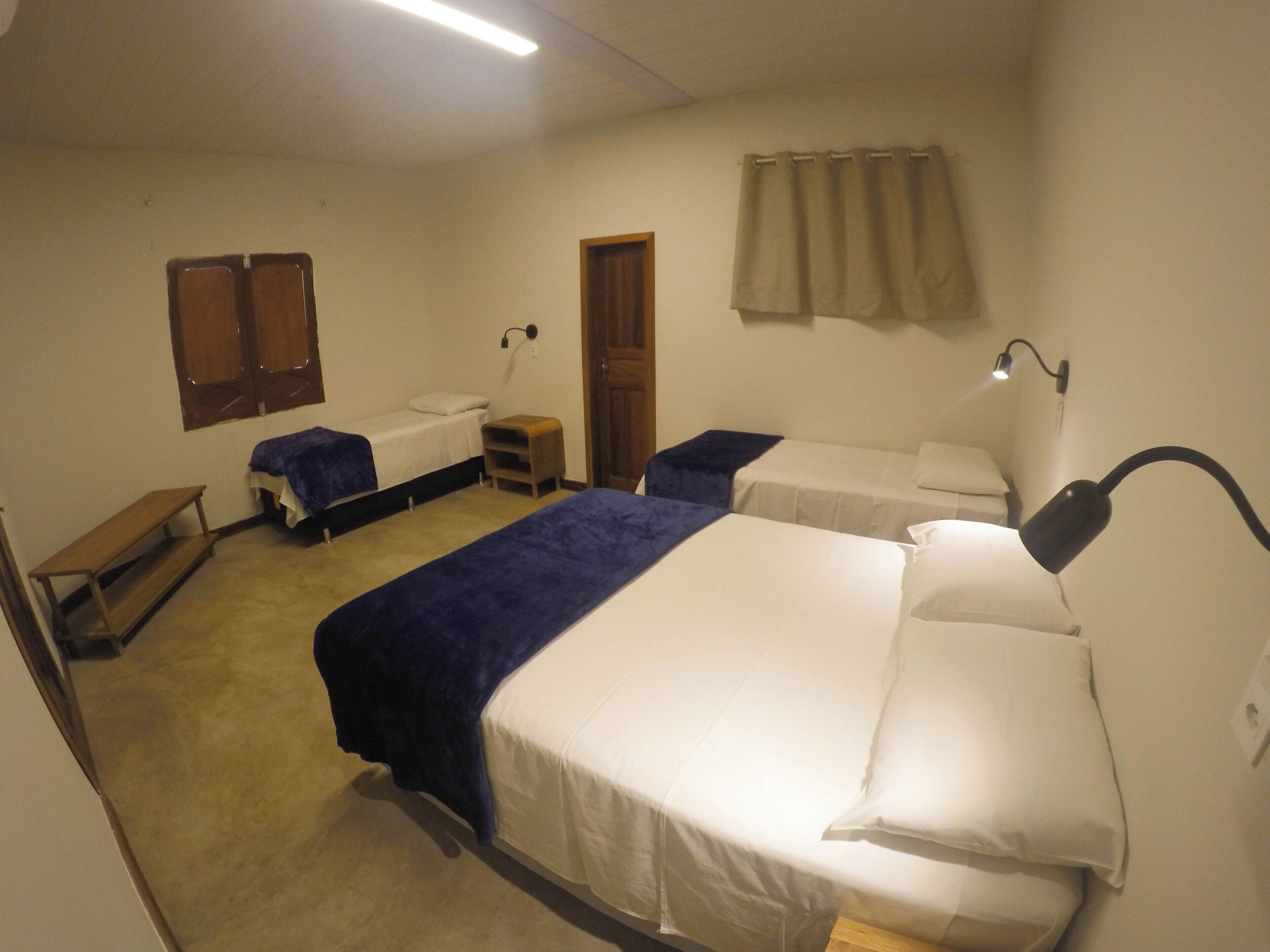 Quarto com duas camas, um banco, uma escrivaninha, uma porta e uma cortina.