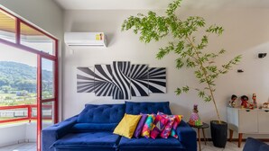 Sala moderna com um sofá azul, almofadas coloridas, um grande quadro abstrato, uma planta de bambu e vista para uma paisagem verde através de uma janela ampla.