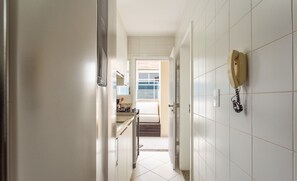 Um corredor estreito com paredes revestidas de azulejos brancos, uma área de cozinha com fogão e bancada visíveis e um telefone fixado na parede.