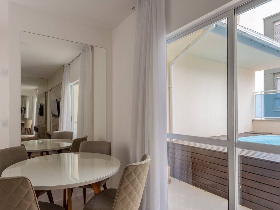 Uma sala de jantar moderna com mesa de vidro e cadeiras acolchoadas, um espelho grande e vista para uma piscina e área externa.