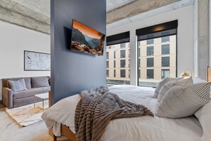 Um quarto moderno com uma cama grande, um sofá cinzento, uma mesinha e uma janela ampla com vista para a cidade.