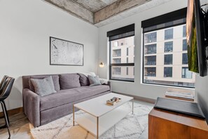 Uma sala de estar moderna com um sofá cinzento, uma mesa de centro branca e uma janela grande com vista para edifícios.