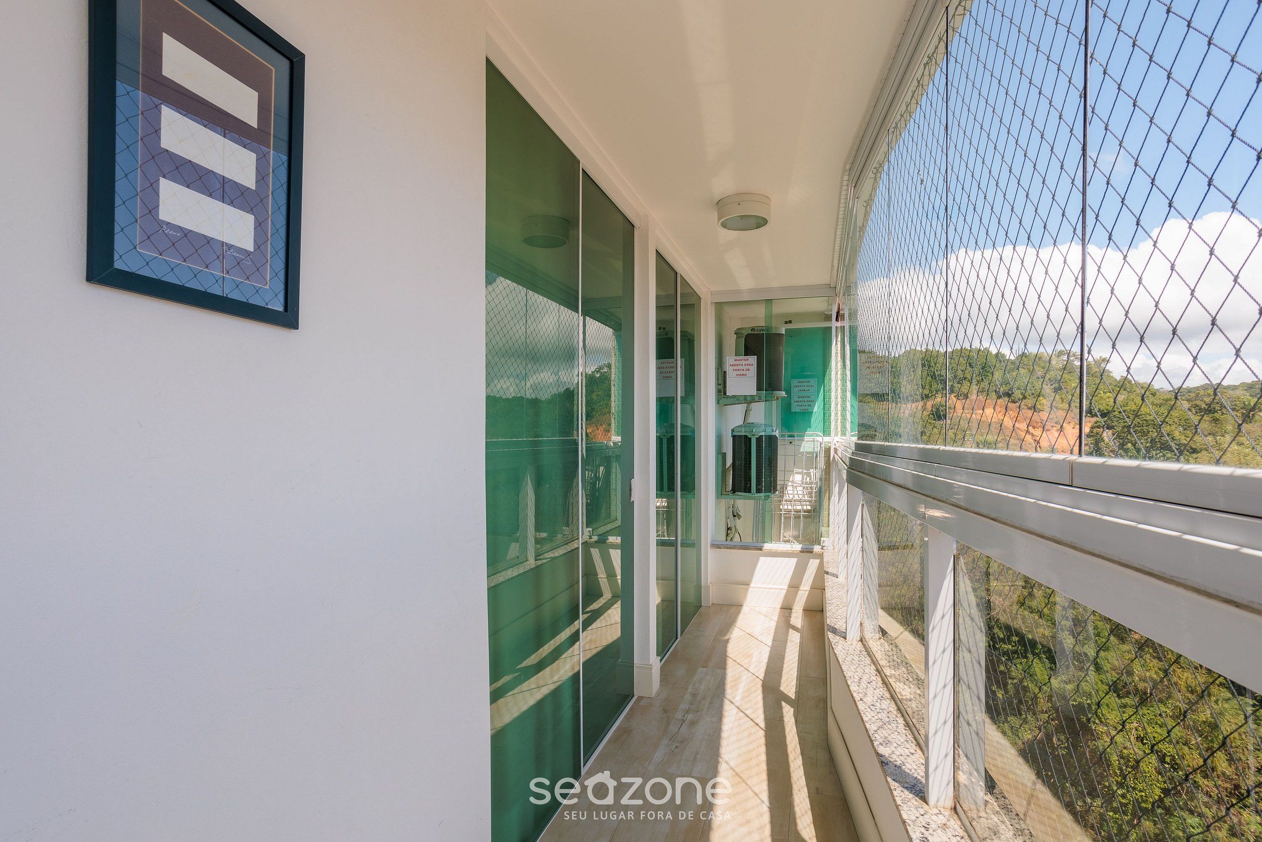 Uma varanda com porta de vidro que dá para um quarto, um quadro na parede e vista para o verde lá fora.