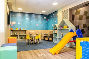 Uma sala de brinquedos infantil com um escorrega, cadeiras coloridas e uma parede azul com objetos suspensos.