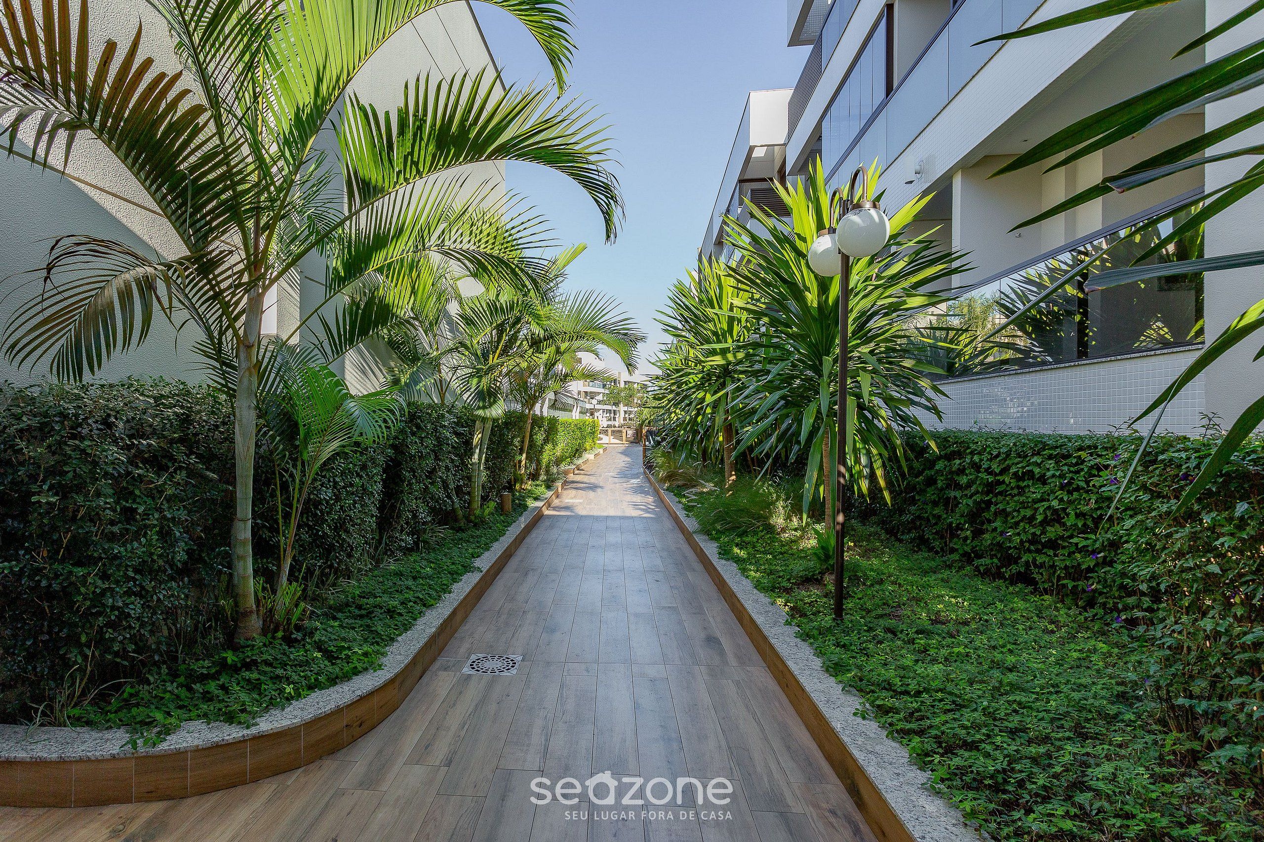 Um caminho ladeado por vegetação exuberante e edifícios modernos, conduzindo a uma entrada distante.