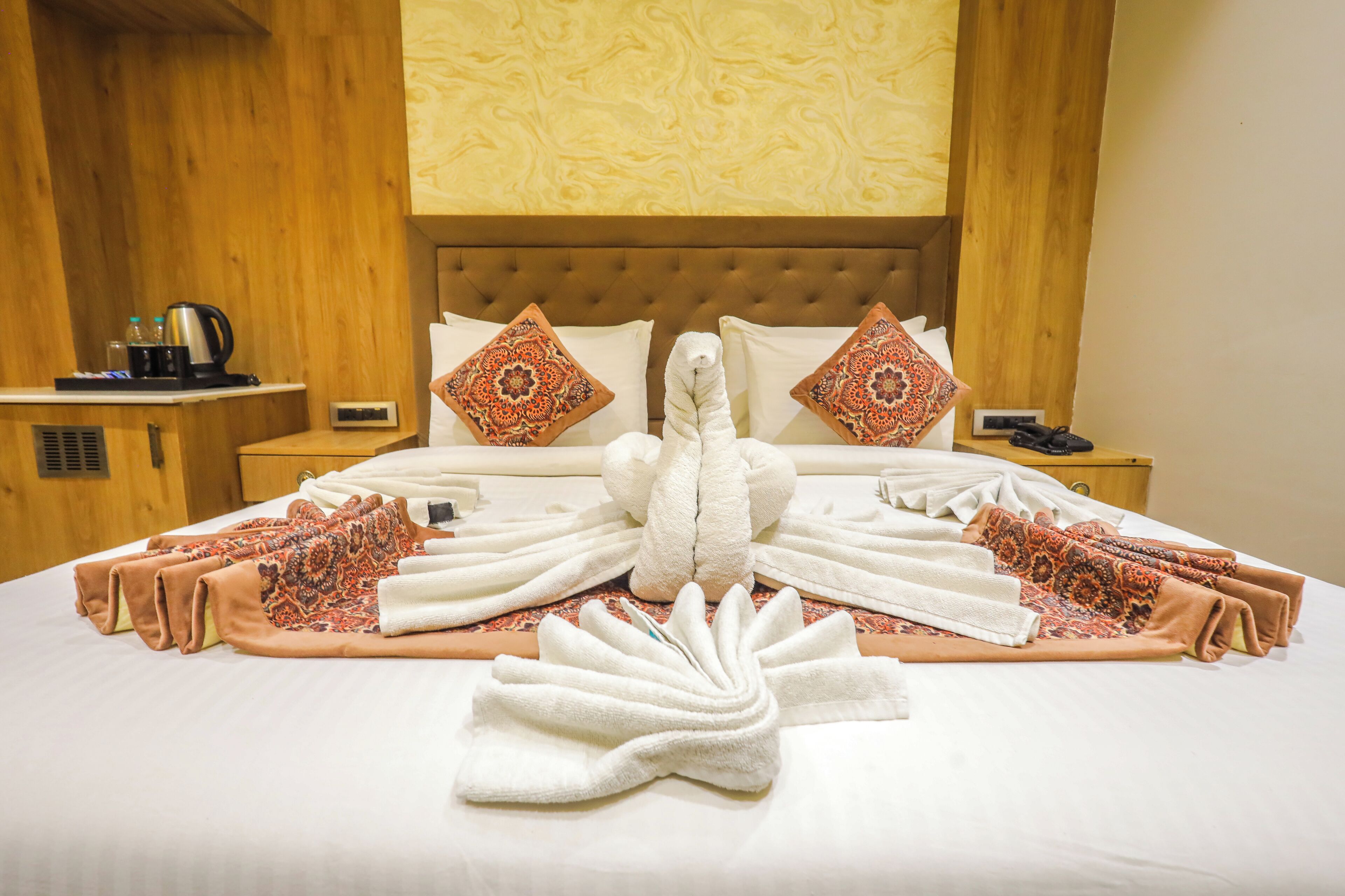 Cama bem arrumada com toalhas dispostas em formato de cisne, cabeceira de madeira, almofadas estampadas e uma pequena mesa com chaleira e xícaras.