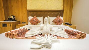 Cama bem arrumada com toalhas dispostas em formato de cisne, cabeceira de madeira, almofadas estampadas e uma pequena mesa com chaleira e xícaras.