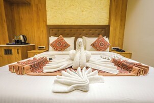Cama bem arrumada com toalhas dispostas em formato de cisne, cabeceira de madeira, almofadas estampadas e uma pequena mesa com chaleira e xícaras.