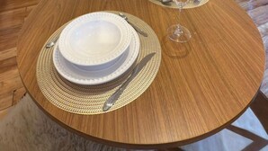 Uma área de refeições com uma mesa redonda de madeira posta para dois, uma geladeira branca, micro-ondas e uma pia ao fundo.