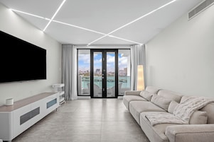 Sala de estar moderna com televisão de tela plana, sofá, mesa de centro e porta de vidro deslizante para uma varanda com vista para a cidade.