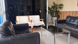 Uma sala de estar moderna com um sofá de couro preto, um sofá branco e uma mesa de centro de vidro. Há uma grande planta em vaso e uma parede de destaque em madeira.
