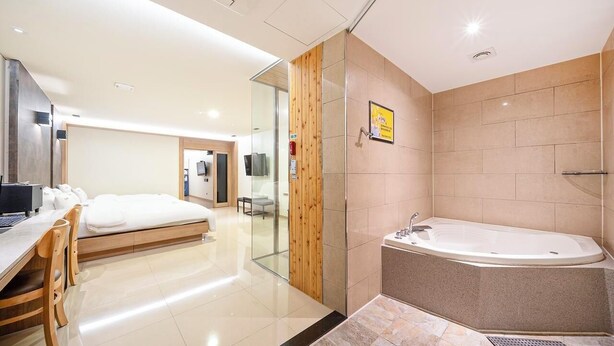 Quarto de hotel moderno com uma cama, uma escrivaninha, uma banheira grande e um separador de vidro que divide o quarto do banheiro.